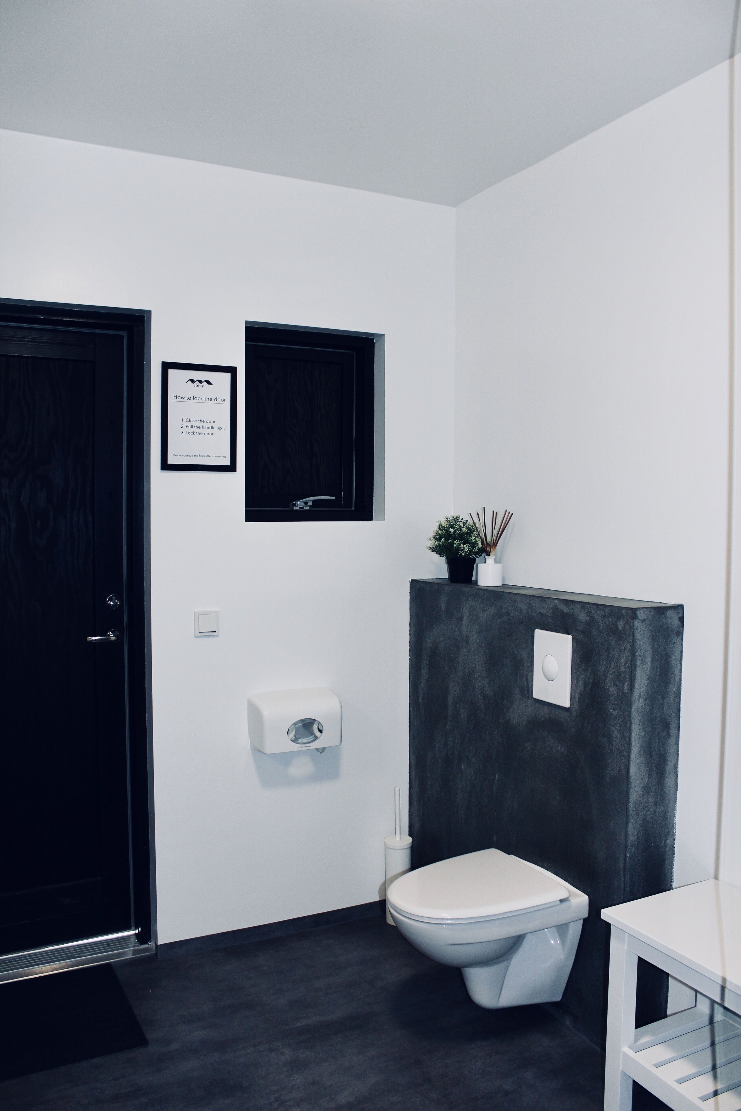 Uma casa de banho moderna com sanita, secador de mãos, porta preta e uma pequena planta num móvel.