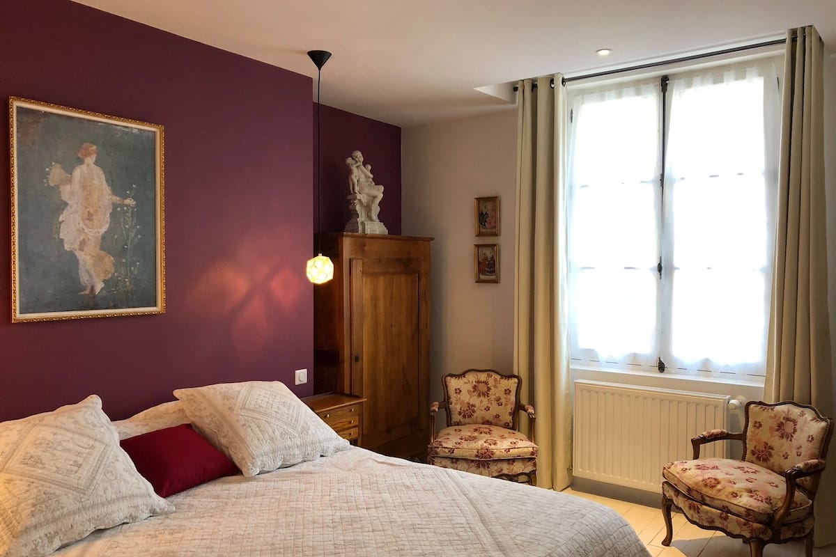 Quarto com uma cama grande, duas cadeiras, um armário de madeira, um quadro e uma janela com cortinas.