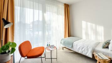 Quarto de hotel com cama, uma cadeira laranja, uma mesinha com abajur e uma planta.