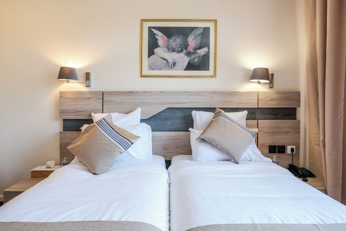 Quarto de hotel com duas camas, cabeceira, abajures, uma moldura na parede e um telefone na mesinha de cabeceira.