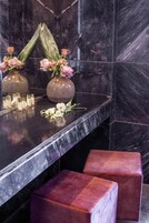 Uma bancada de mármore com dois vasos de flores cor-de-rosa e um pequeno buquê, um conjunto de velas e dois taboretes vermelhos acolchoados.