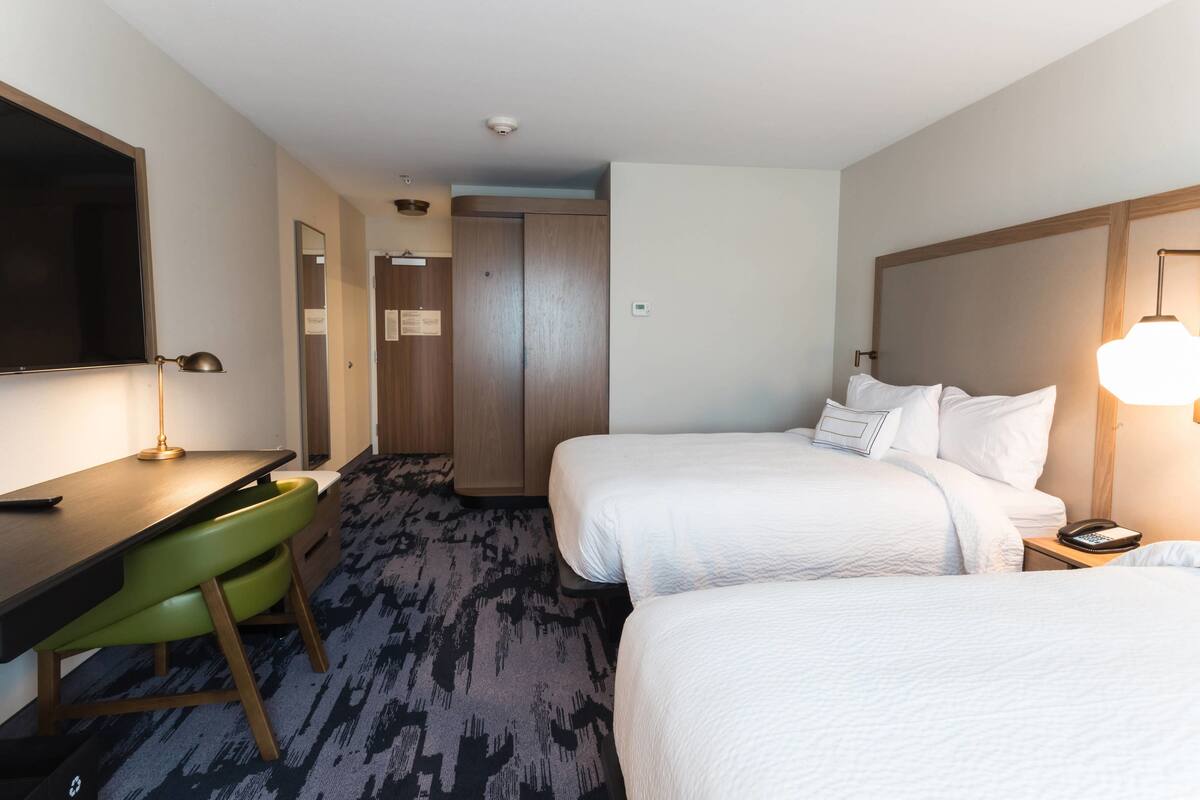 Quarto de hotel com duas camas, uma escrivaninha, uma cadeira, uma TV e um abajur.