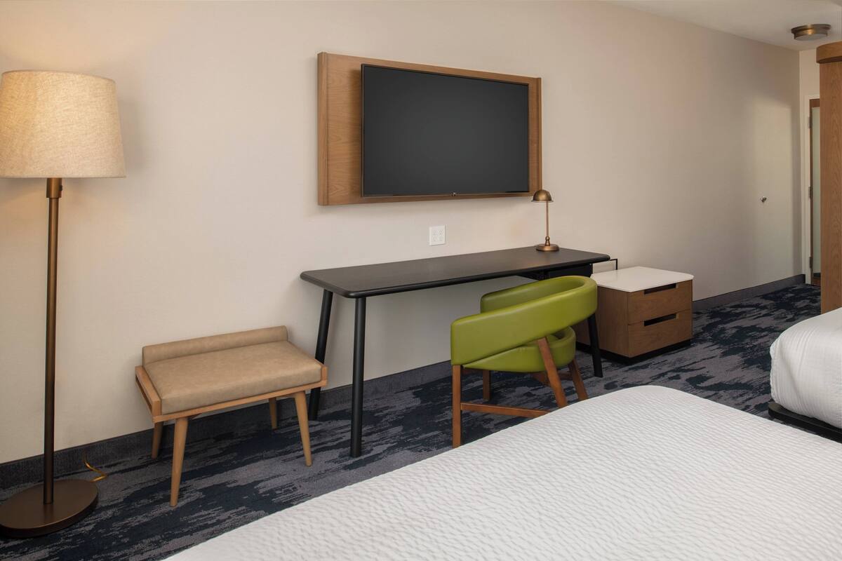 Quarto de hotel com cama, escrivaninha, cadeira, abajur e televisão.