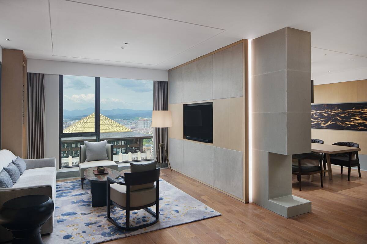 Quarto de hotel moderno com uma janela grande, televisão de tela plana, sofá, mesa de centro e área de refeições.