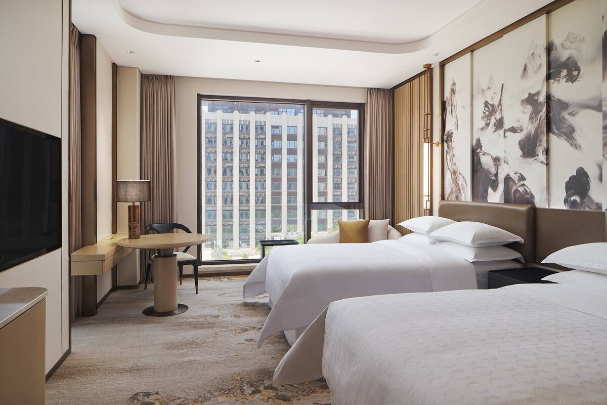 Quarto de hotel com duas camas, uma escrivaninha, uma cadeira, televisão e uma janela grande com vista para a cidade.