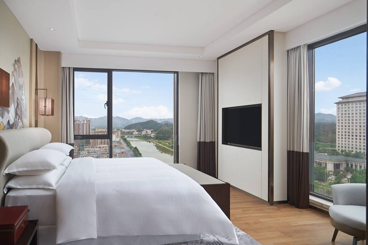 Quarto de hotel moderno com uma cama grande, televisão de tela plana e vista para a cidade através de janelas amplas.
