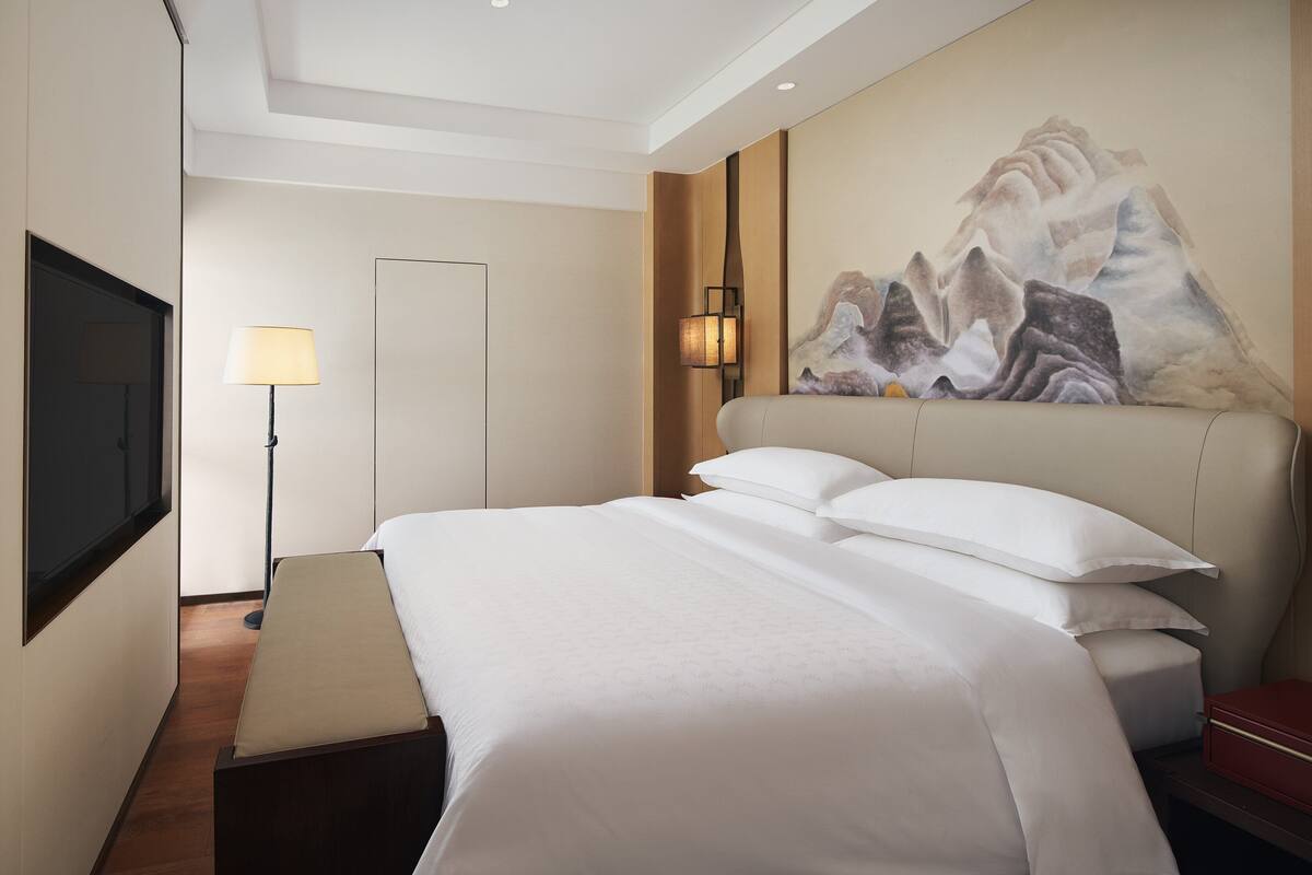 Quarto de hotel com uma cama grande, um quadro de montanhas na parede, uma mesa de cabeceira e um abajur.
