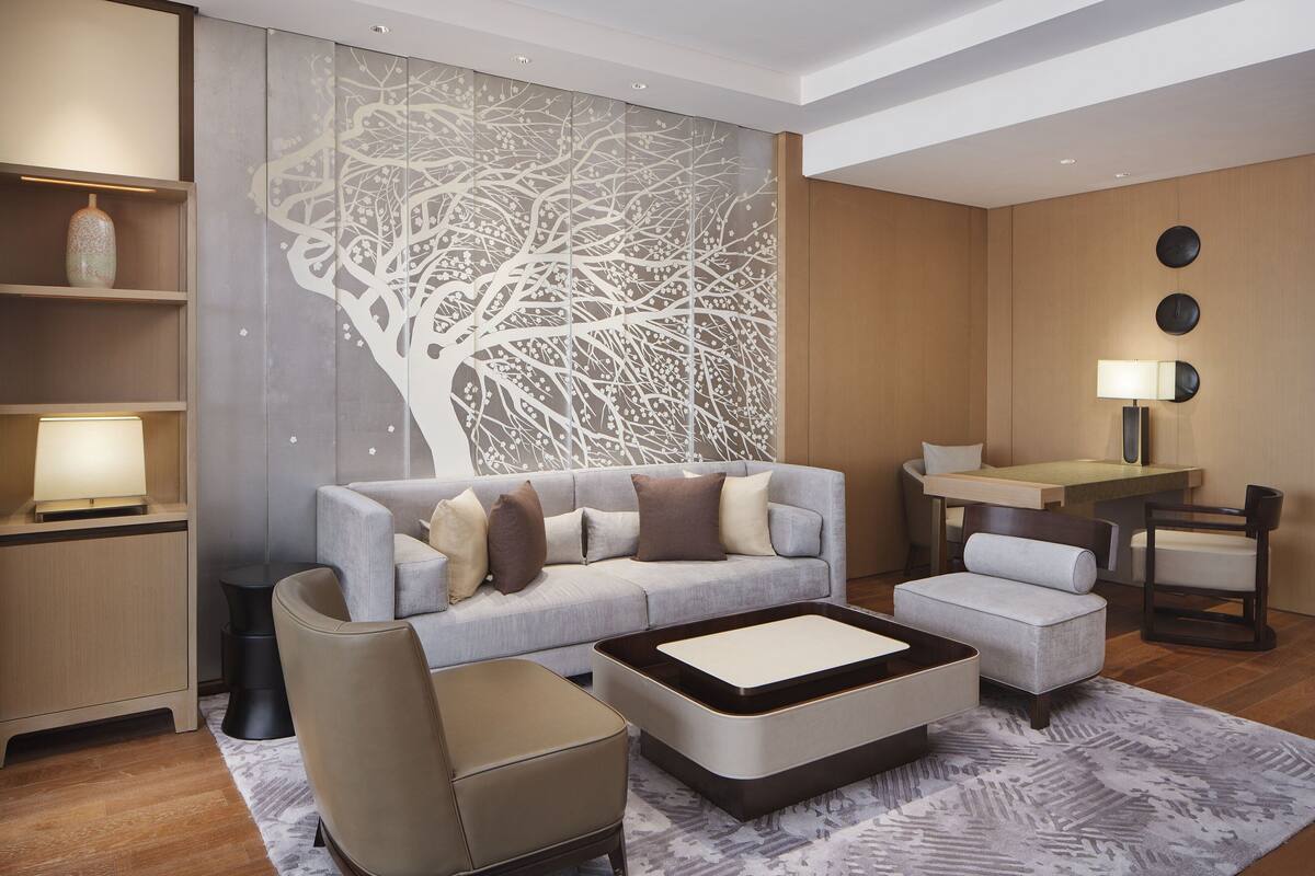 Uma sala de estar moderna com um sofá cinzento, um banquinho pequeno, uma mesa de centro e um mural decorativo com uma árvore na parede.