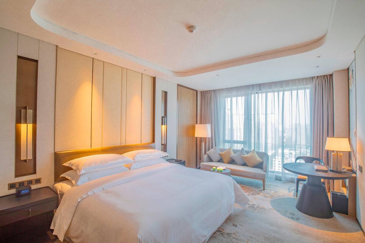 Quarto de hotel com uma cama grande, um sofá, uma mesa de jantar e vista para a cidade por grandes janelas.