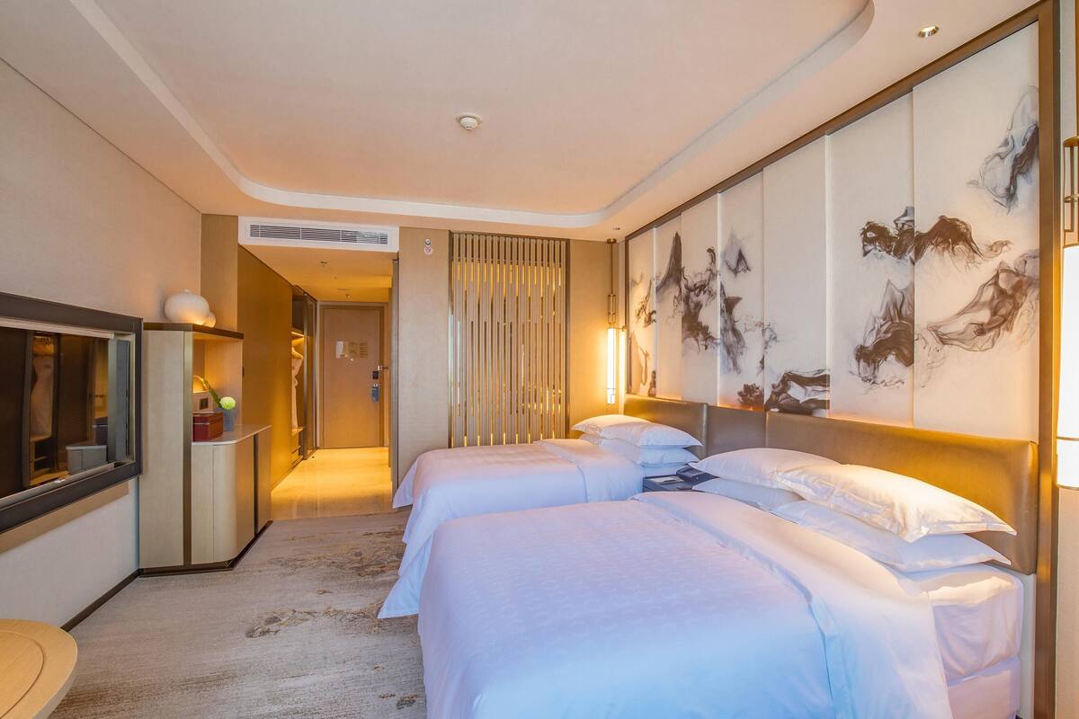 Quarto de hotel com duas camas, televisão, escrivaninha com luminária e obras de arte na parede.