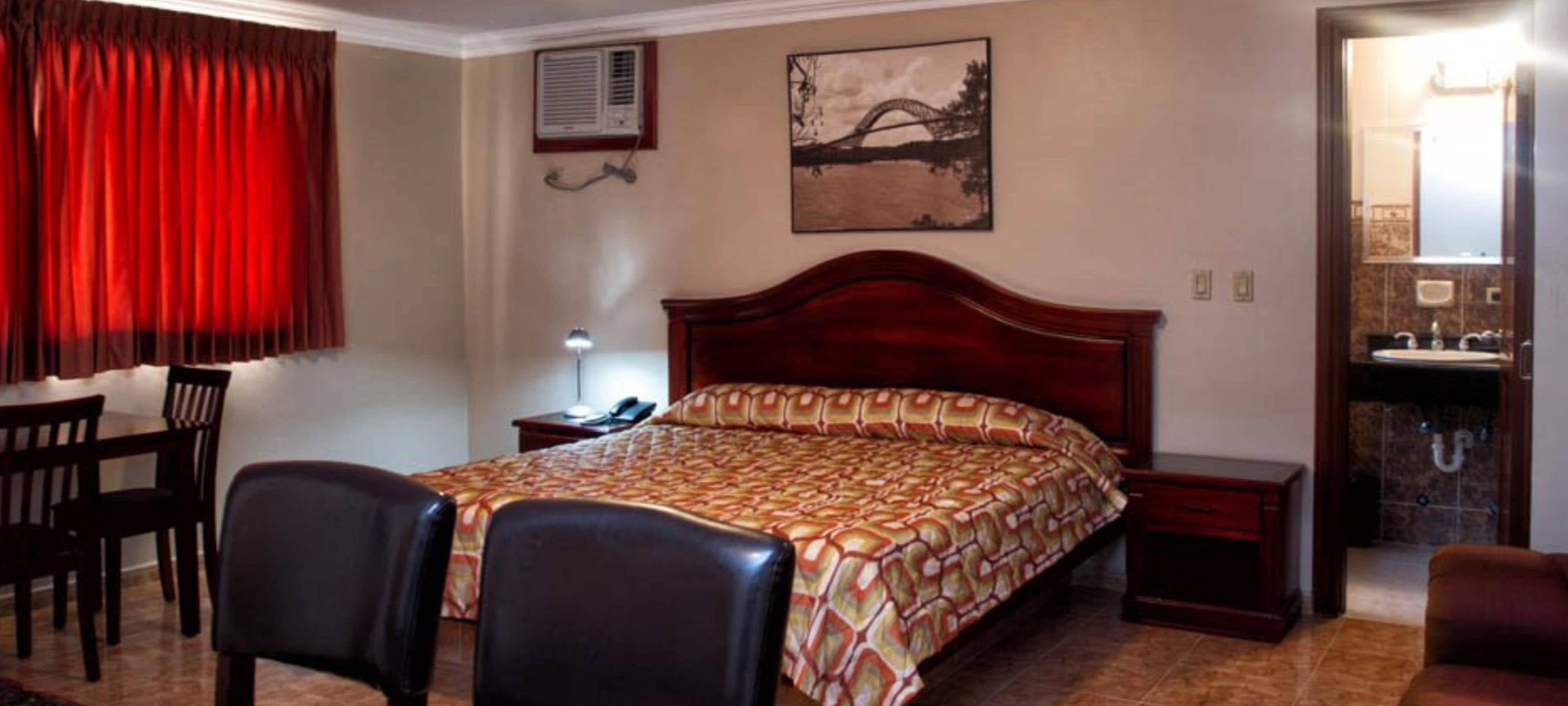 Quarto de hotel com cama, escrivaninha, duas cadeiras, mesa de cabeceira, luminária, um quadro na parede e banheiro visível através de uma porta aberta.
