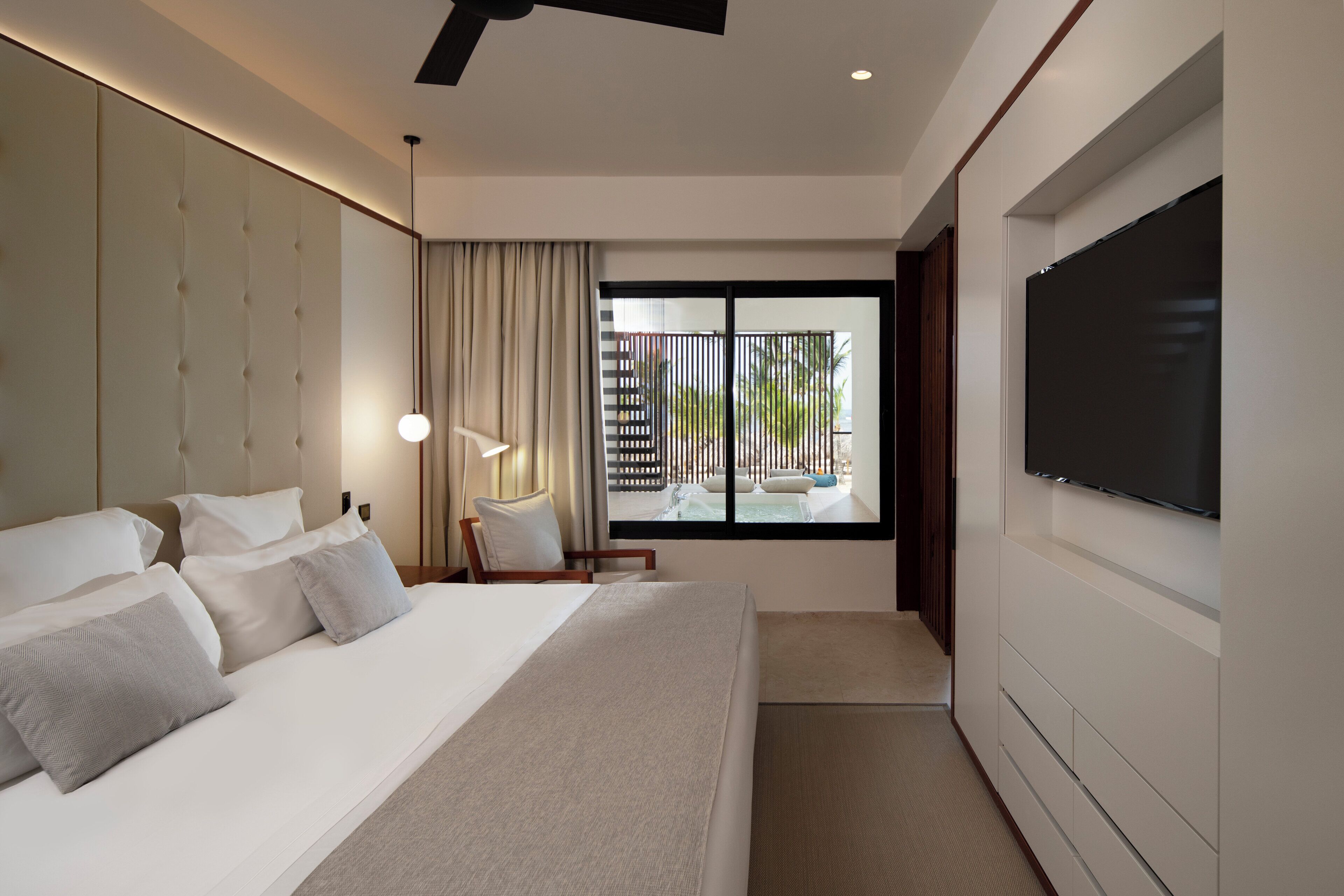 Quarto de hotel moderno com uma cama grande, televisão de tela plana, ventilador de teto e vista para o exterior por uma porta de correr.