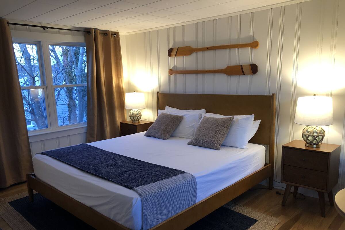 Cama bem arrumada com lençóis brancos e manta azul, acompanhada por duas mesas de cabeceira com abajures, cabeceira de madeira e vista para árvores pela janela.