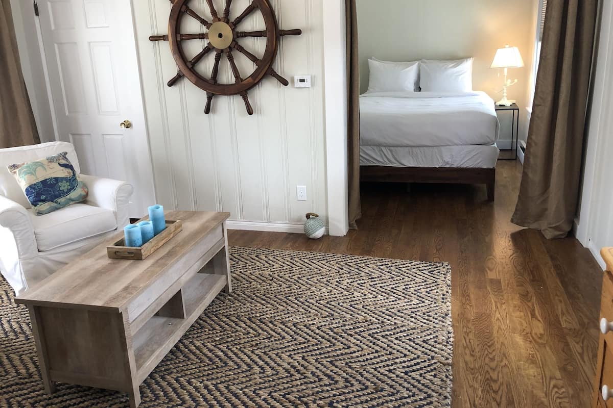 Quarto com mesa de centro de madeira, sofá branco com almofada estampada, decoração de parede em formato de leme de navio, cama com roupa de cama branca e tapete estampado.