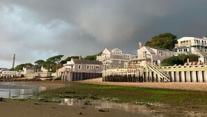 Uma área costeira com um cais, casas à beira-mar e um farol ao fundo.