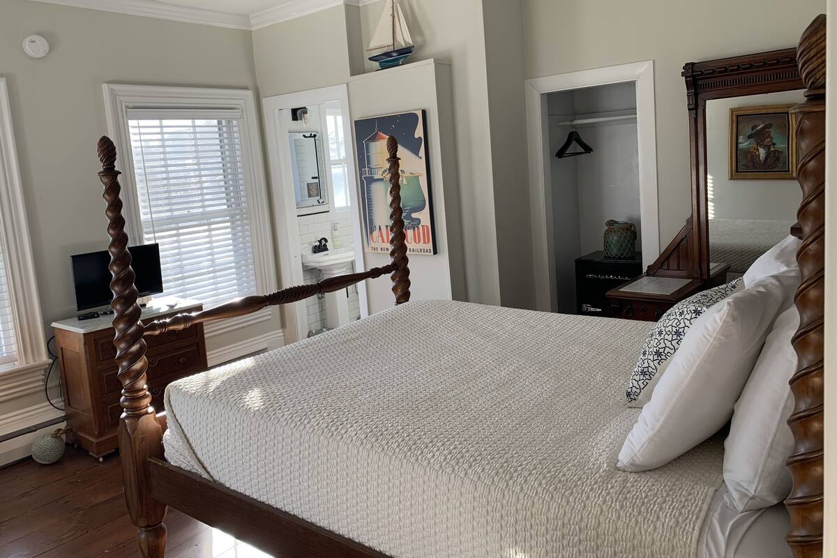 Quarto com cama de dossel, penteadeira de madeira, televisão e banheiro visível por uma porta aberta.