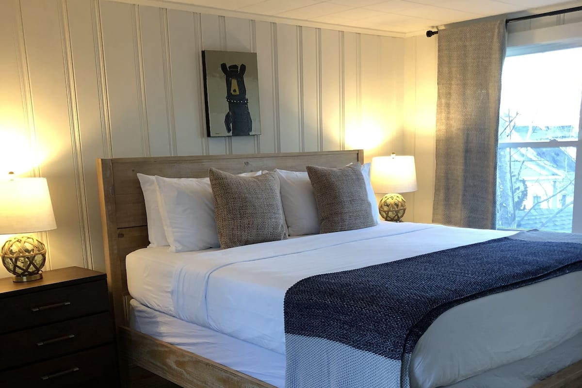 Cama bem arrumada com roupa de cama branca e cinza, cabeceira de madeira e manta azul escura. Criado-mudo com luminária e objeto decorativo ao lado da cama. O quarto tem piso de madeira e uma grande janela com cortinas.