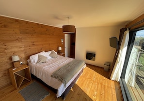 Um quarto com piso de madeira, uma cama com travesseiros, uma mesa de cabeceira com abajur, um lustre pendente e uma janela com cortinas.