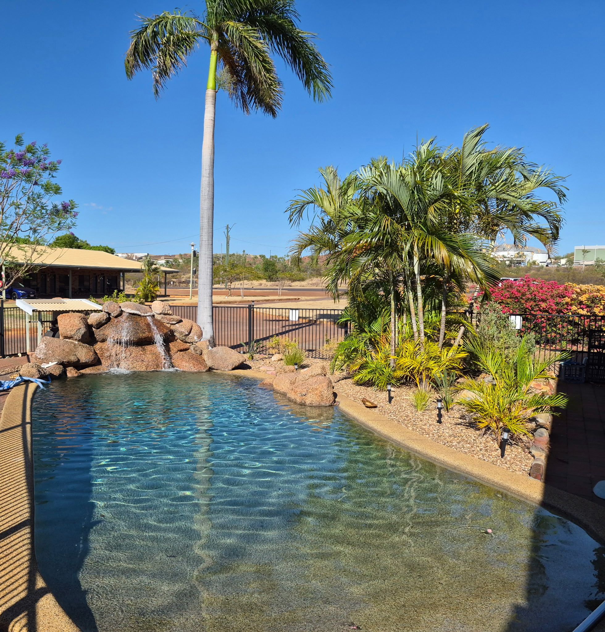 Piscina cristalina com deck de madeira, cercada por palmeiras e paisagem rochosa.