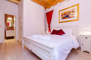 Quarto com cama grande, roupa de cama branca, cortina vermelha, mesa de cabeceira com abajur e um quadro na parede.