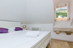 Um quarto com uma cama, roupa de cama branca, uma pequena mesa de cabeceira com uma lâmpada, uma janela com vista para um edifício e vegetação, e um parapeito de pedra abaixo da janela.