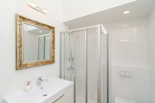 Uma casa de banho moderna com uma toucador branca, um espelho com moldura dourada, cabine de duche em vidro e parede revestida a azulejos brancos.