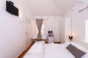 Quarto de hotel com cama, escrivaninha com luminária, televisão e janela com cortinas.