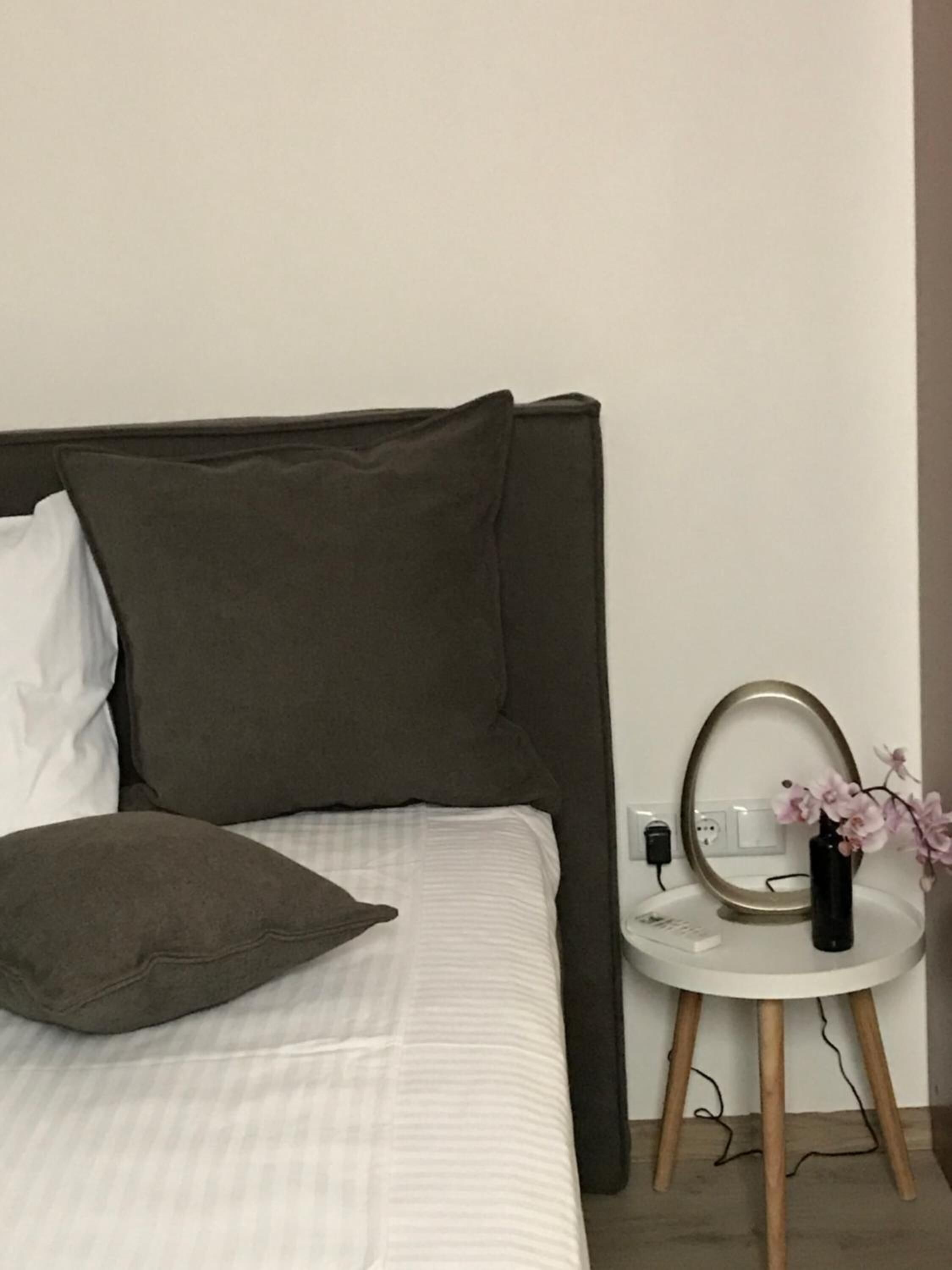 Cama bem arrumada com colcha listrada em branco e cinza, cabeceira marrom escura e um único travesseiro marrom escuro. Uma pequena mesa de cabeceira branca com um espelho redondo e um vaso de flores é visível.