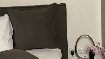 Cama bem arrumada com colcha listrada em branco e cinza, cabeceira marrom escura e um único travesseiro marrom escuro. Uma pequena mesa de cabeceira branca com um espelho redondo e um vaso de flores é visível.