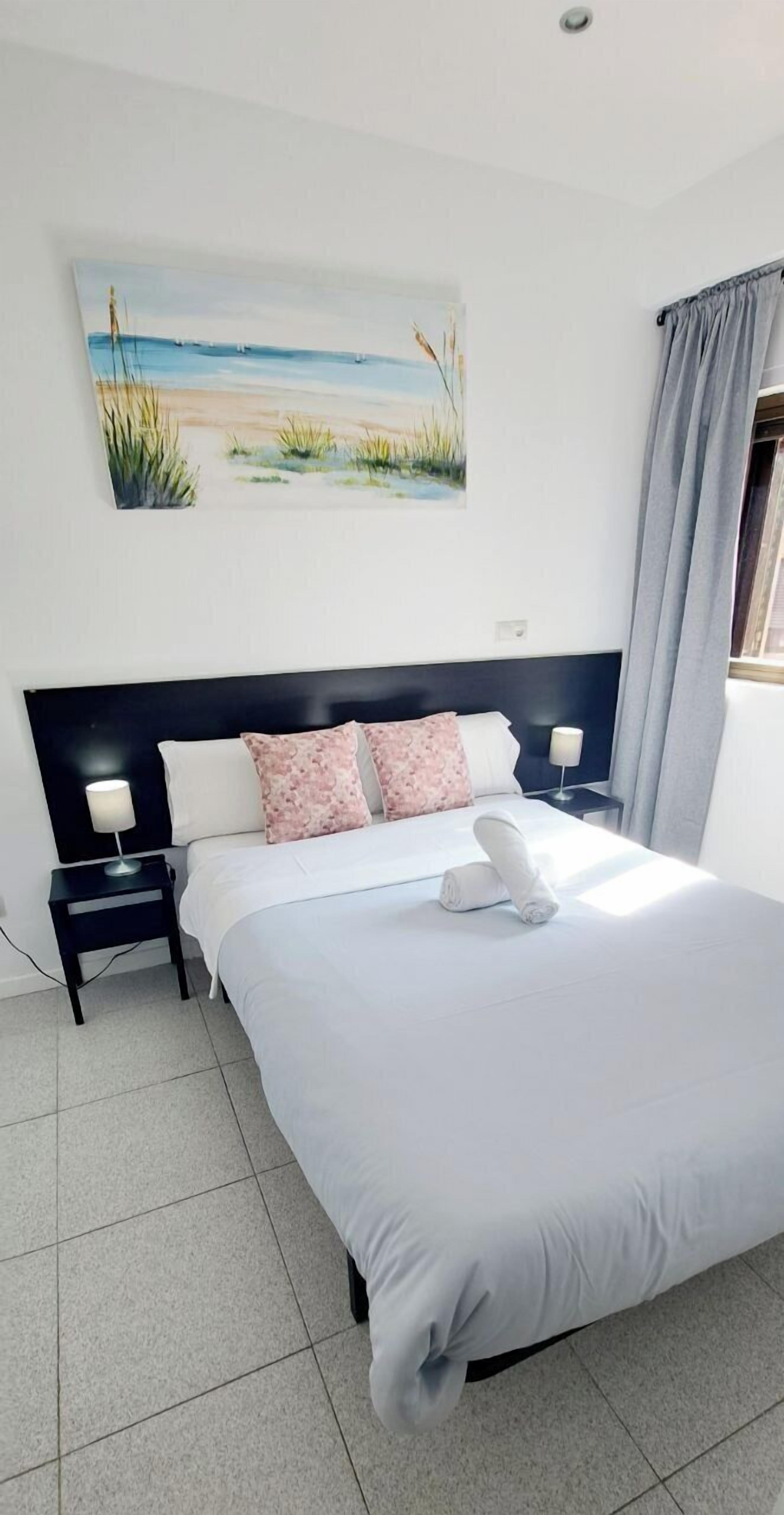Uma cama bem arrumada com edredom branco e almofadas florais cor-de-rosa, uma mesa de cabeceira com abajur e um quadro com uma paisagem de praia na parede.