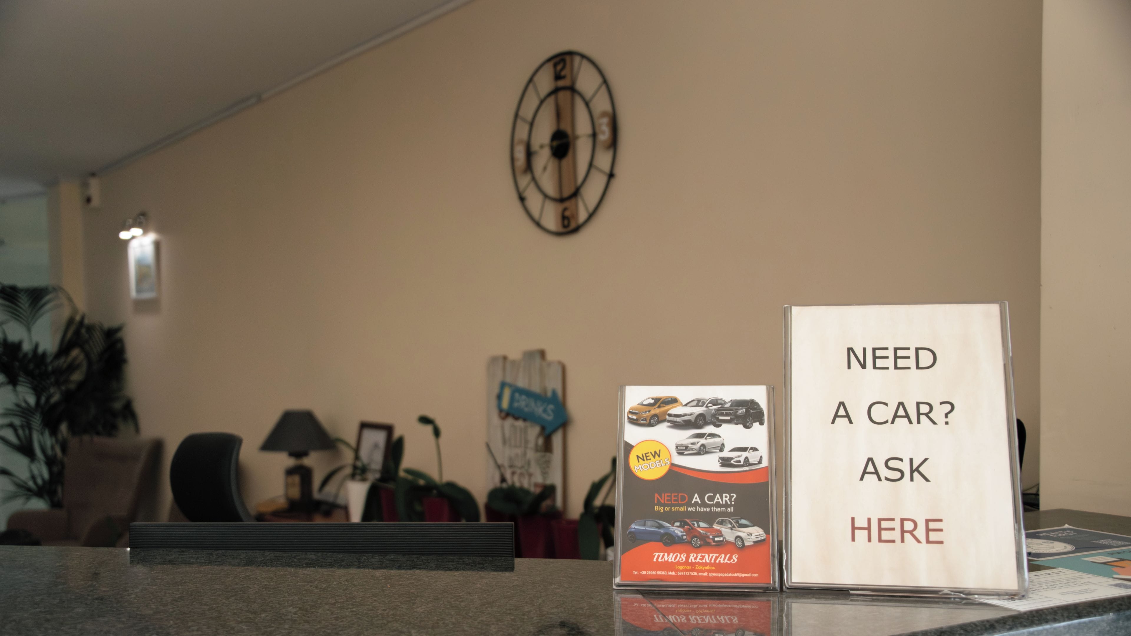 Balcão de recepção com um relógio na parede, um aviso oferecendo aluguel de carros e um aviso que diz 'PRECISA DE UM CARRO? PERGUNTE AQUI'.