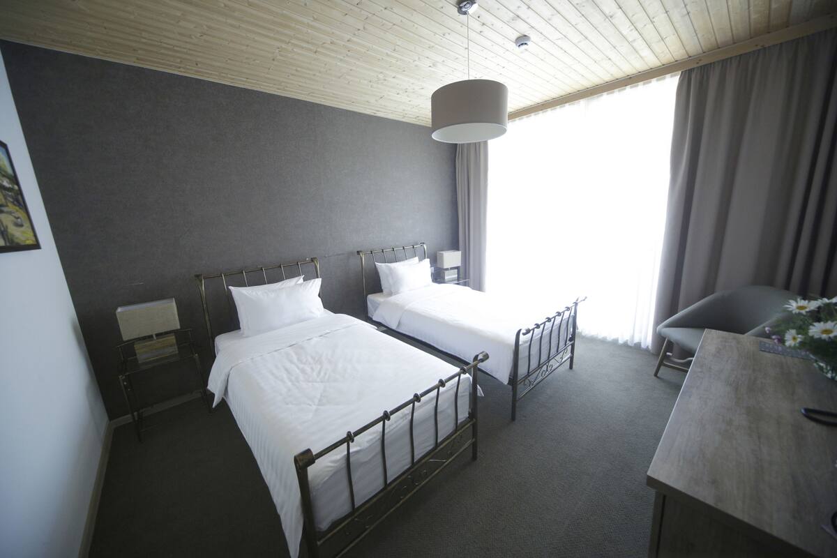 Quarto de hotel com duas camas de solteiro, uma escrivaninha, uma cadeira, um abajur e uma janela grande com cortinas.