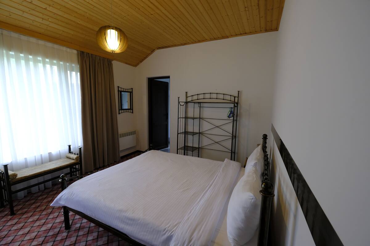 Um quarto com cama, um banco, uma janela com cortinas e uma porta.