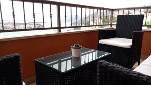 Uma varanda com móveis de vime, uma mesa de vidro e um vaso com planta.