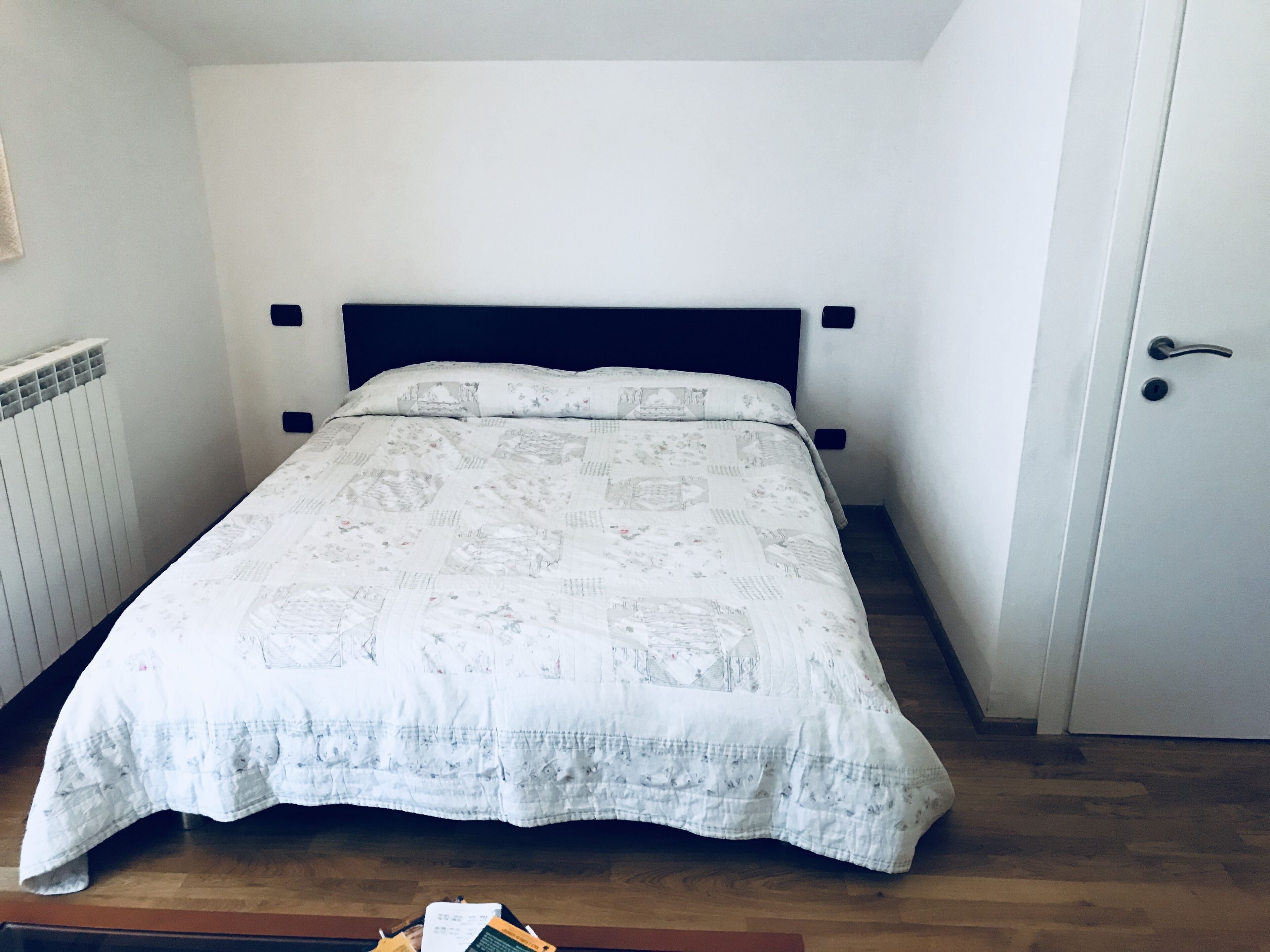 Quarto pequeno com uma cama de solteiro, cabeceira escura, paredes brancas e piso de madeira.