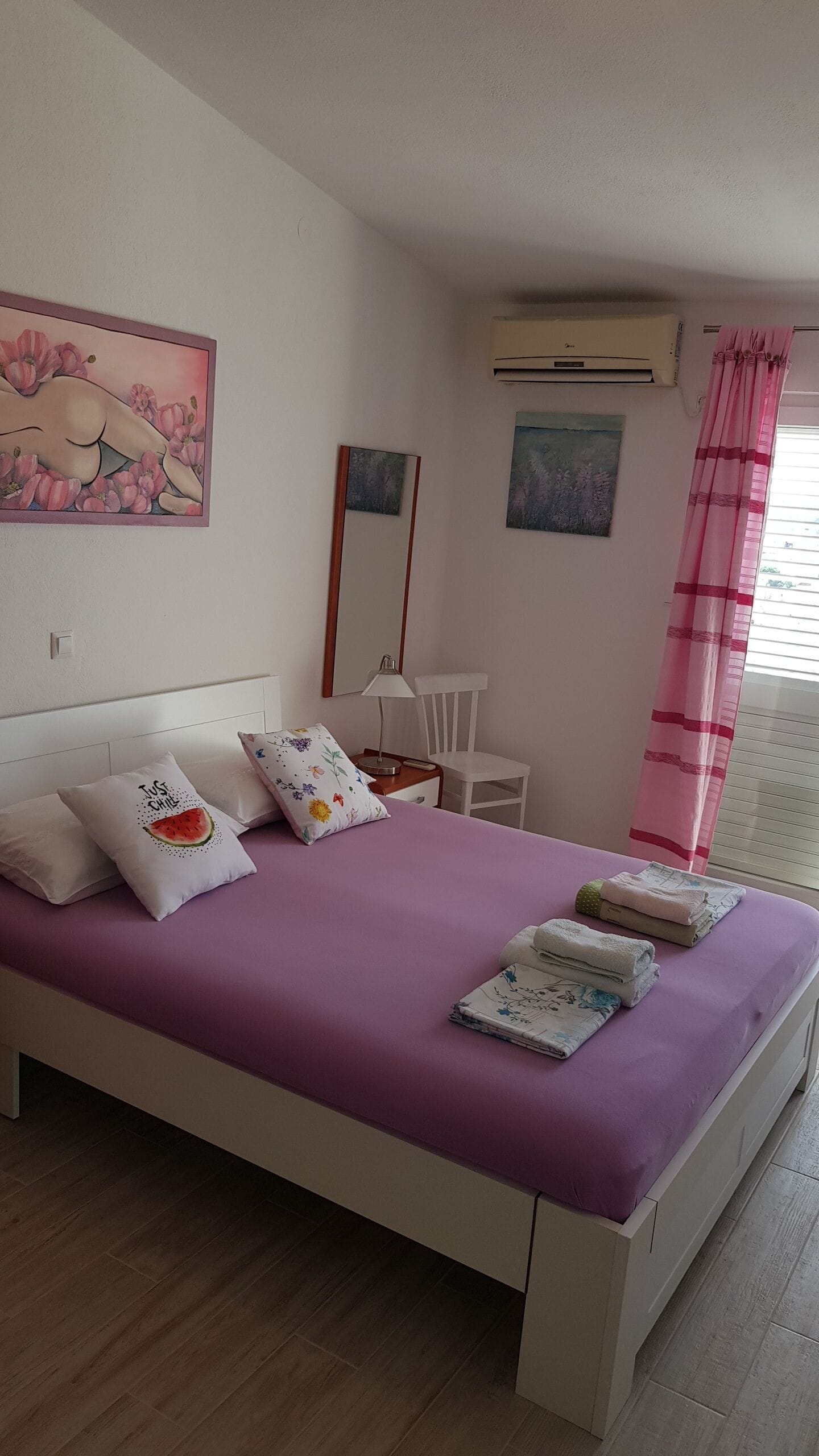 Quarto com cama, almofadas, mesa de cabeceira, cadeira, abajur, ar condicionado e janela com cortinas.