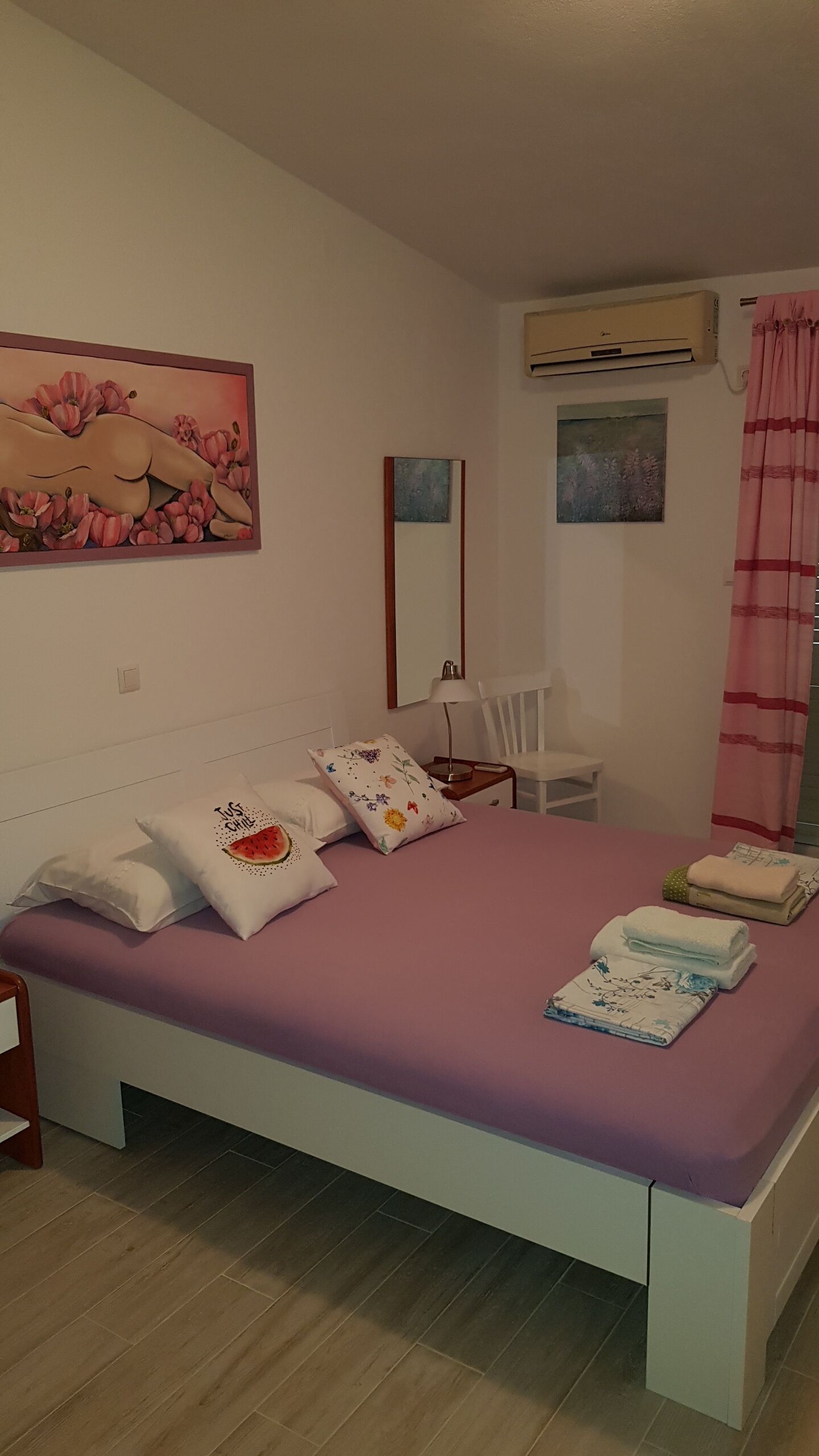 Quarto com cama, travesseiros, poltrona, mesa de cabeceira, luminária, ar-condicionado e um quadro na parede.