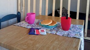 Uma mesa de madeira com toalha de mesa florida, duas chávenas cor-de-rosa, um prato com dois brioches e uma embalagem de snacks.