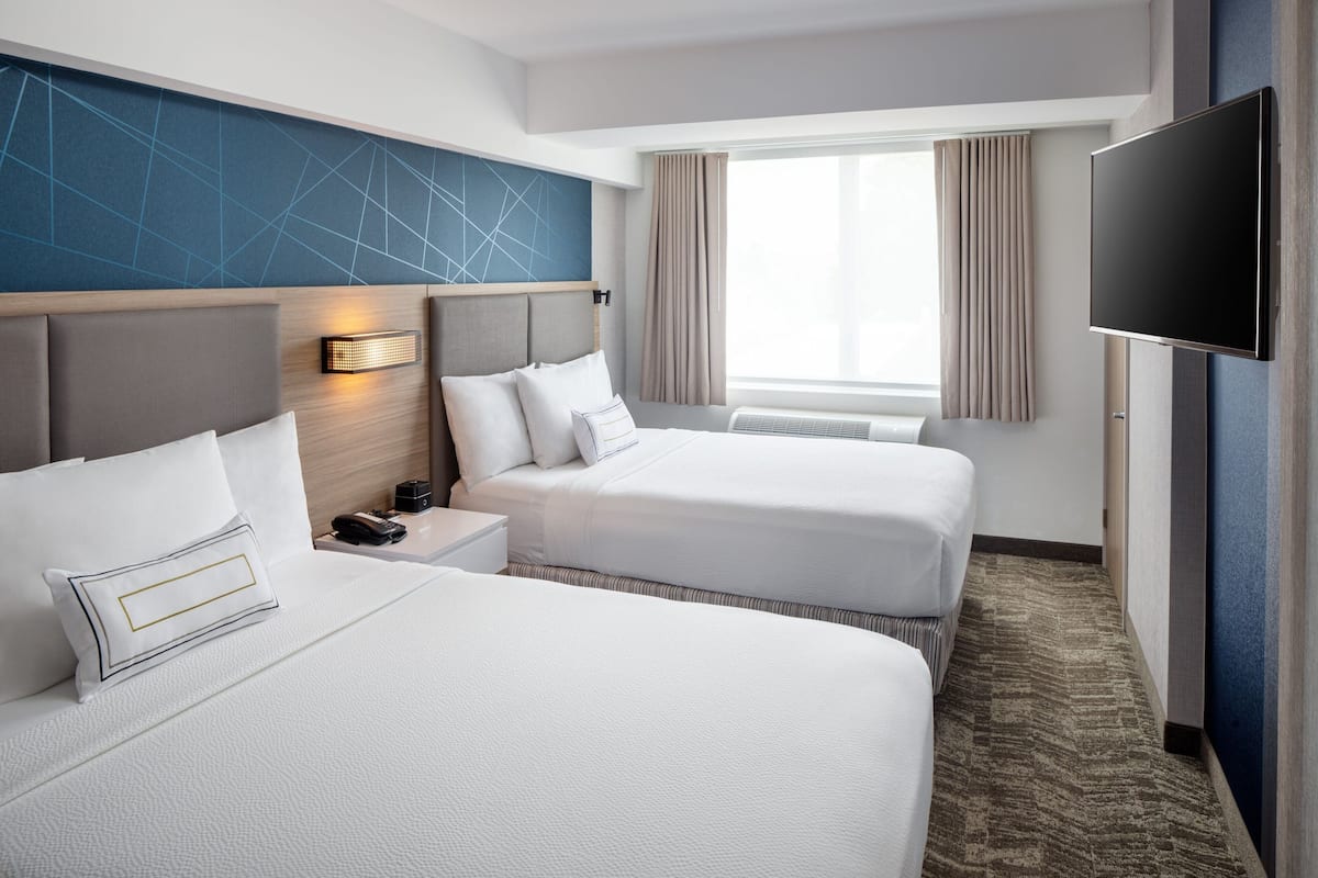 Quarto de hotel com duas camas, televisão, mesa de cabeceira com abajur, janela com cortinas e televisão montada na parede.