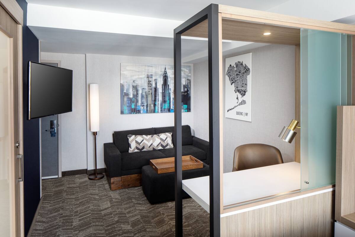 Quarto de hotel moderno com televisão de tela plana, sofá preto, mesa de cabeceira de madeira, divisória de vidro e uma pintura com vista para a cidade.