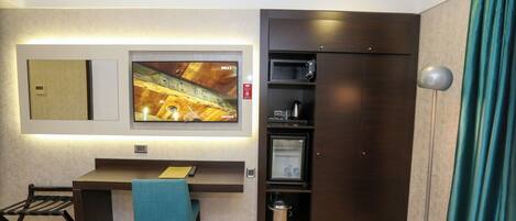 Quarto de hotel com uma cama, escrivaninha, cadeira, televisão, micro-ondas e guarda-roupa.