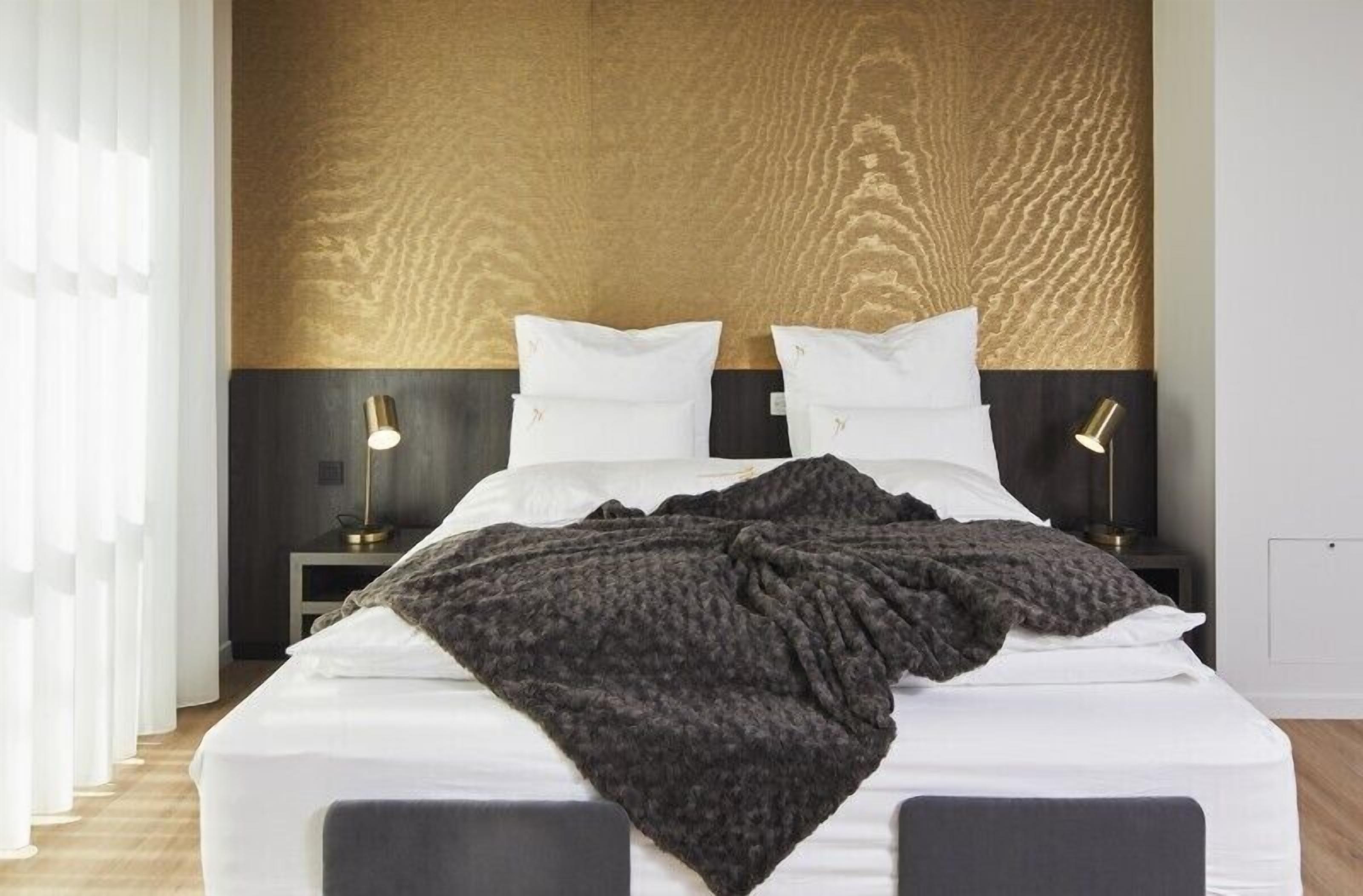 Cama bem arrumada com roupa de cama branca e manta cinza escura. Duas mesas de cabeceira com abajures e uma parede texturizada ao fundo da cama.