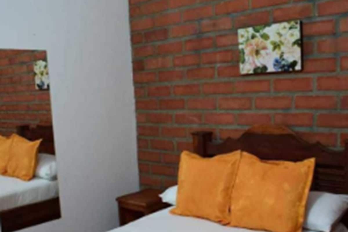 Quarto de hotel com parede de tijolos, cama com colcha laranja, mesa de cabeceira e um quadro na parede.