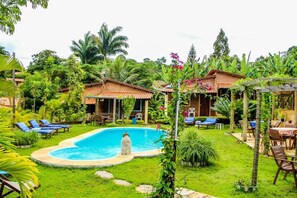 Uma piscina rodeada por vegetação exuberante, com uma pequena estrutura de pedra no centro. Há cadeiras de descanso e uma área de refeições com mesas e cadeiras.