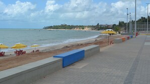 Um calçadão à beira-mar com um banco de concreto, guarda-sóis amarelos e cadeiras vermelhas na orla.