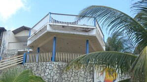 Um edifício de dois andares com varanda, paredes de pedra e uma palmeira em primeiro plano.