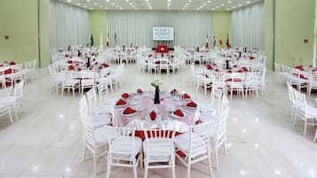 Uma grande sala de conferências com várias mesas redondas, cada uma com cadeiras brancas e toalhas de mesa vermelhas.