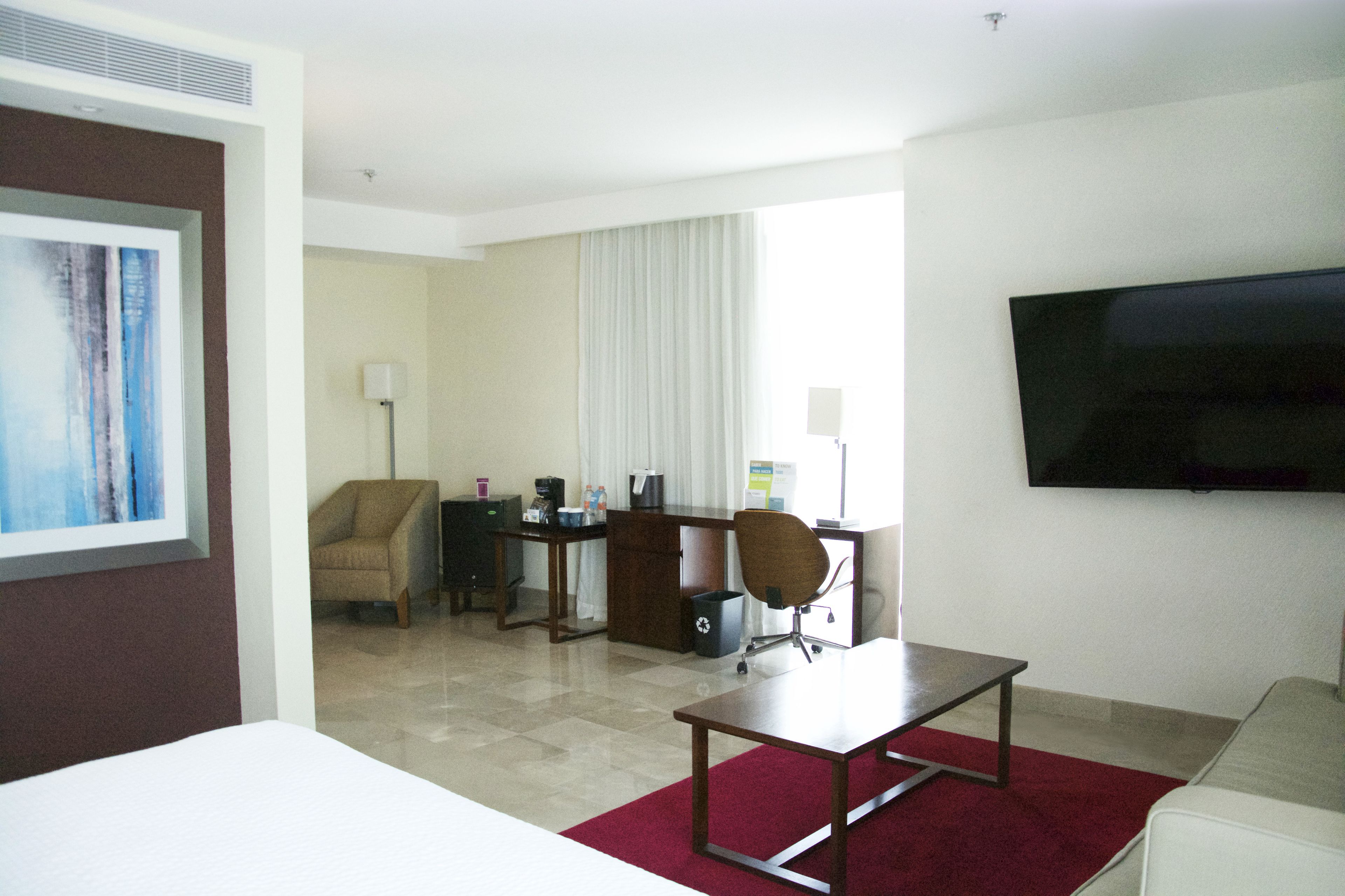 Quarto de hotel com cama, escrivaninha, cadeira, televisão e um quadro na parede.