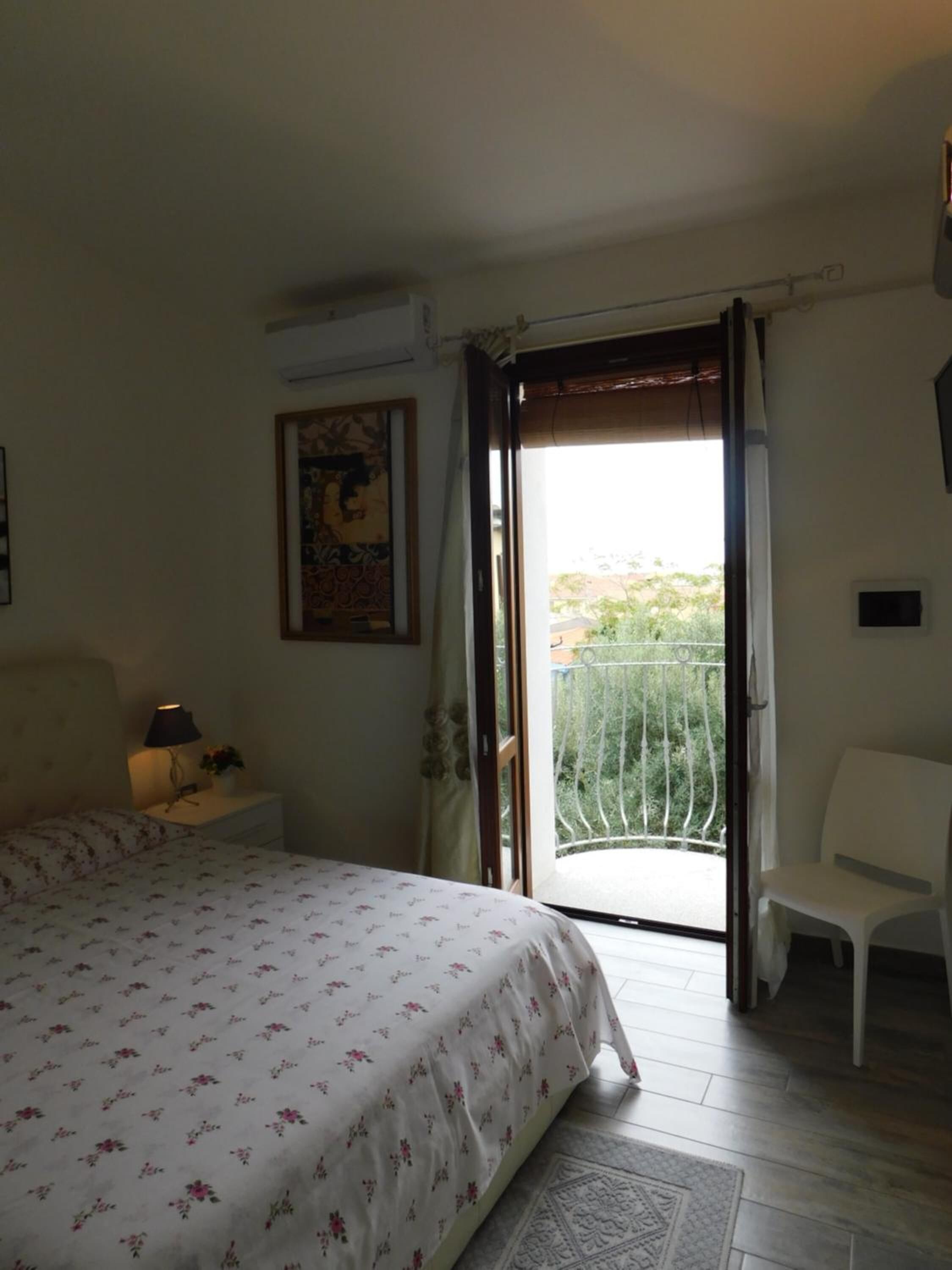 Quarto com cama, mesa de cabeceira com luminária, uma cadeira e varanda com vista para o verde.