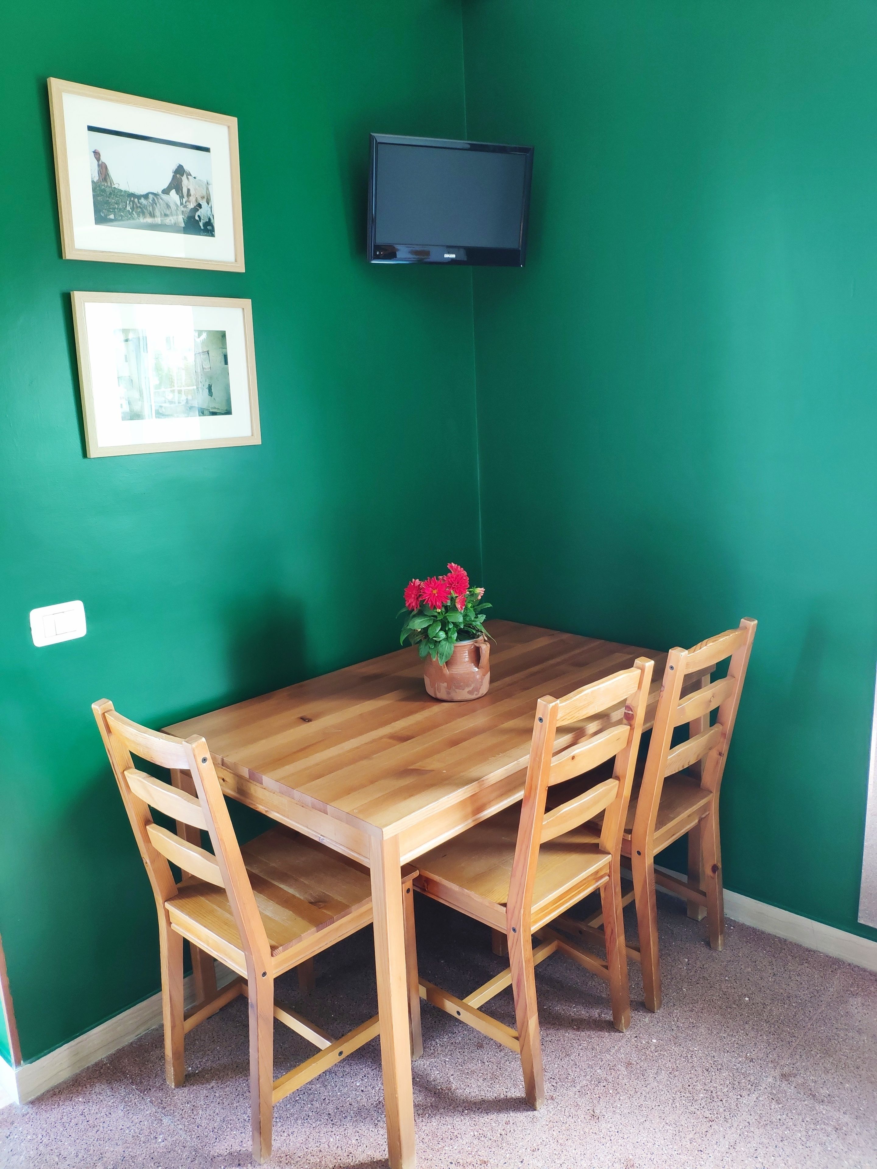 Uma área de refeições com uma mesa de madeira e quatro cadeiras, uma televisão fixada na parede e duas molduras com imagens.